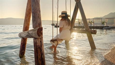 긴 흰색 드레스를 입은 젊은 아름 다운 여성이 일몰 광선이 비추는 바다 해변에서 밧줄 그네에서 휴식을 취하고 있습니다 관광 여름철 휴가 및 해변에서의 휴가 30 34세에