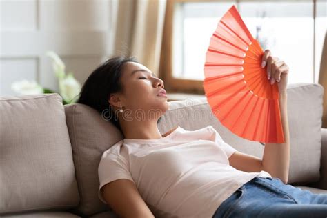Overheated Exhausted Asian Woman Waving Paper Fan Sitting On Couch