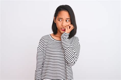 줄무늬 티셔츠를 입은 젊은 중국 여성은 손톱을 물고 손톱을 물고 손에 스트레스와 긴장 찾고 고립 된 흰색 배경 위에 서있다 불안