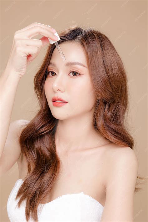 얼굴에 한국식 화장을 하고 피펫을 사용하여 고립된 베이지색 배경의 피부에 세럼을 바르는 젊은 아시아 미인 여성 곱슬머리 미용
