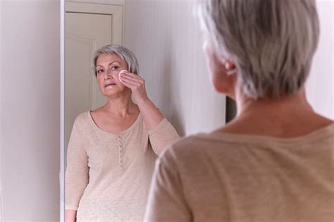 캐주얼 한 옷을 입고 짧은 회색 머리를 가진 노인 백인 여성은 얼굴의 피부를 케어합니다 55 59세에 대한 스톡 사진 및 기타 이미지 55 59세 가정 생활 거울