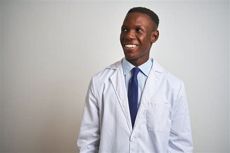 고립된 흰색 배경 위에 코트를 입고 있는 젊은 아프리카 계 미국인 의사가 얼굴에 미소를 지으며 옆으로 바라보고 자연스러운 표정 자신감 있게 웃고 있습니다 감정에 대한 스톡