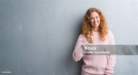 젊은 빨강 머리 여자 회색 그런 지 벽 입고 위에 분홍색 스웨터 행복 엄지손가락 손으로 제스처를 하 고 승인 성공을 보여주는