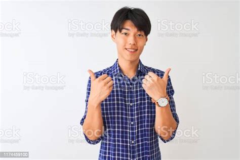 고립 된 흰색 배경 성공 표지판 위에 서있는 캐주얼 한 파란색 셔츠를 입은 젊은 중국 남자는 손으로 긍정적 인 제스처를하고 엄지 손가락으로 미소와 행복을 합니다 쾌활한