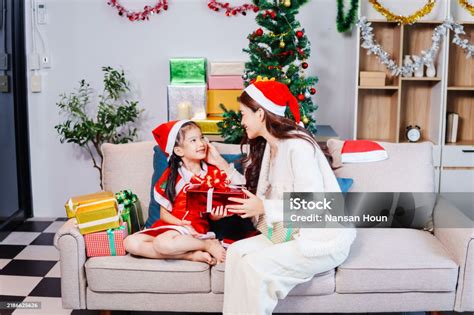 예쁜 엄마와 귀여운 딸이 집의 소파에 앉아 선물 상자를 들고 기쁜 듯이 웃고 있다 생일 케이크와 장식된 크리스마스 트리가 축제 분위기를 더합니다 12월에 대한 스톡 사진 및