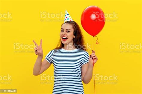 생일 모자를 쓴 예쁜 젊은 여성은 노란색 배경에 빨간색 팽창 헬륨 풍선을 들고 평화 제스처를 취합니다 휴일과 파티 개념 20