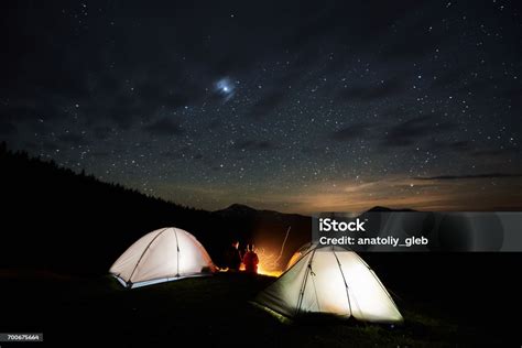 밤 산에 캠핑입니다 로맨틱 커플 관광객의 후면 보기 밤에 별이 빛나는 하늘 아래 두 개의 조명된 텐트 근처 캠프 파이어에서