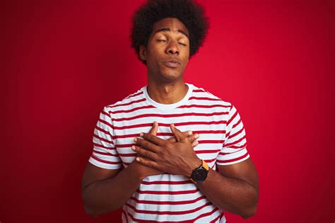 고립 된 빨간 배경 위에 줄무늬 티셔츠를 입은 젊은 아프리카 계 미국인 남성은 닫힌 눈과 얼굴에 감사하는 제스처로 가슴에 손을 대고 미소짓고 있습니다 건강 개념 T 셔츠에