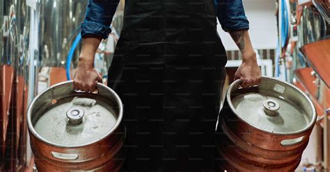 앞치마를 입은 맥주 생산 공장의 남성 노동자가 거대한 강철 탱크의 줄을 따라 카메라 앞에서 움직이면서 두 개의 수조를 들고 있다 사진 Unsplash의 증류소 이미지