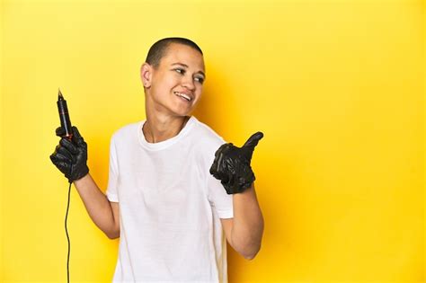 Mulher tatuadora em estúdio cabeça raspada fundo amarelo aponta