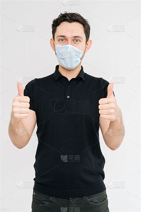 工作室拍摄的乐观英俊的男子戴着医用口罩，黑色t恤显示拇指向上的手势