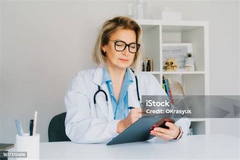 자신감 넘치는 성숙한 중년 여성 의사 의료 간호사 흰 코트를 입은 여성 의사가 청진기를 들고 병원 사무실의 작업장에 앉아 약속을 지키는 동안 메모를 하고 있다 45 49세에