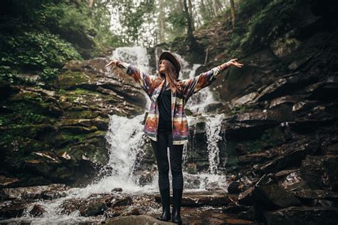 여행과 방 랑 벽 개념입니다 폭포 산의 숲에 서 있는 배낭과 모자에 세련 된 Hipster 소녀 숲에서 손을 키우는 여행자 여자