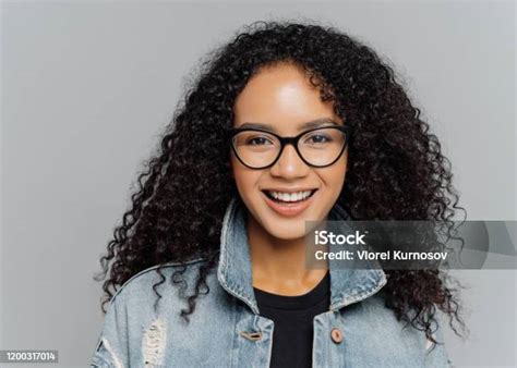 곱슬 머리를 가진 기쁜 여자 광학 안경을 착용 데님 재킷 회색 배경에 고립 된 카메라에 똑바로 보이는 완성 된 작업에 만족 사랑스러운 대화를 즐긴다 얼굴 표정 T 셔츠에