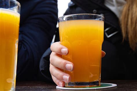 흐린 오렌지 주스 또는 여과되지 않은 음료 한 잔을 들고 있는 손을 어두운 나무 테이블 위의 컵받침에 올려놓은 손의 클로즈업 감귤류 과일에 대한 스톡 사진 및 기타 이미지