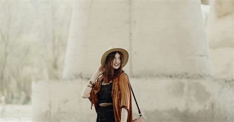 아름 다운 세련 된 힙 스터 여자는 모자 가죽 가방 프린지 판초와 부츠와 함께 걷고 있습니다 다리 아래 해변 근처의 집시 룩을 입은 보호 여행자 소녀 여름 여행