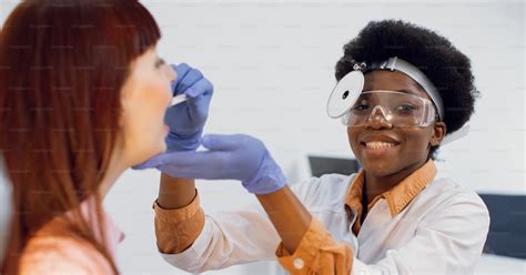 긍정적인 미소를 짓는 젊은 아프리카계 미국인 여의사 이비인후과 의사 유니폼 정면 반사경 및 안경을 착용하고 여성 환자의 목을 확인하고 혀 억제기를 사용합니다 사진