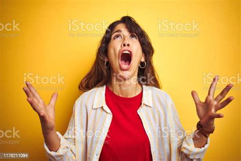 노란색 고립 된 배경에 빨간 티셔츠와 줄무늬 셔츠를 입고 젊은 아름다운 여성은 미친 미친 소리와 공격적인 표정과 팔을 들고 고함
