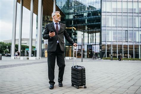 여행 가방과 함께 걷는 공항 근처에서 풀 슈트를 입은 성숙한 사업가 회색 머리 사업가의 전체 길이 비즈니스 웨어 당기는 여행