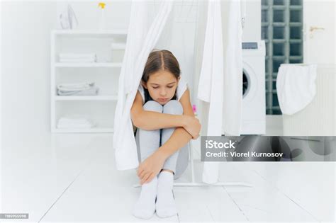 외로운 사려 깊은 슬픈 작은 소녀는 옷 건조기 근처 세탁실에서 바닥에 앉아 바지와 흰색 양말 세탁기 세탁 바구니 및 콘솔을