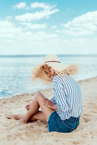 행복 한 흑인 여자 동안 모래 사장에 앉아 카메라를 보고 하는 밀 짚 모자 관광에 대한 스톡 사진 및 기타 이미지 관광