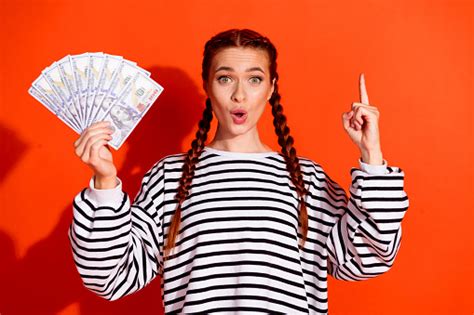 스트라이프 풀오버를 입은 매력적인 빨간 머리 여성이 생생한 주황색 배경을 배경으로 현금을 들고 포즈를 취하며 장난기 넘치는 표정과 흥분을 보여줍니다 땋은 머리에 대한 스톡