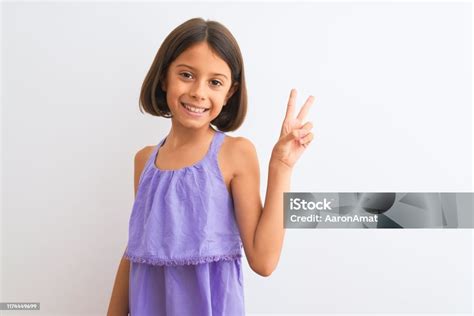 보라색 캐주얼 드레스를 입고 고립 된 흰색 배경 위에 서있는 젊은 아름다운 소녀는 승리의 표지판을하고있는 카메라에서 윙크하는 행복한 얼굴로 미소 짓고 있습니다 2번 갈색