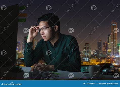 Attractive Young Asian Man Sitting On Desk Table Looking At Laptop Computer In Dark Late Night