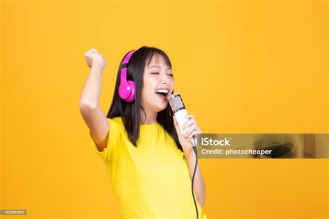 노란색 배경에 노래방 음악을 노래하는 마이크를 들고 노란색 셔츠를 입은 예쁜 아시아 여성 마이크에서 노래를 노래하는 헤드폰으로