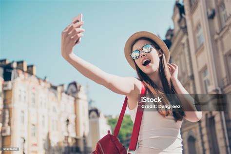 젊은 매력적인 장난 관광이 하고있다 밖에 서 전화에 Selfie를 모자 선글라스 빛 드레스와 빨간 가방 입고 거리에 대한 스톡