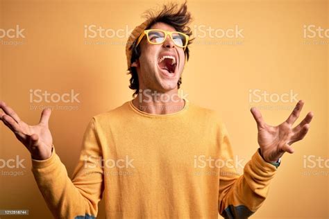 캐주얼 한 티셔츠와 안경을 쓰고 고립 된 노란색 배경에 미친 미친 소리와 공격적인 표정과 팔을 들고 고함을 지르는 젊은 잘생긴
