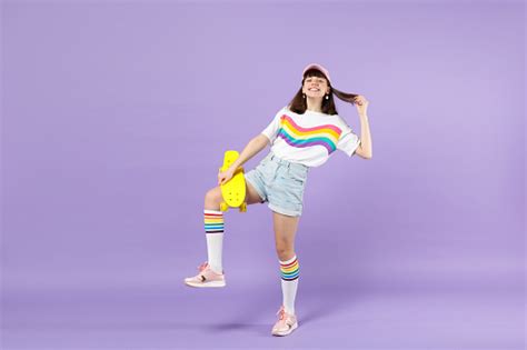 스튜디오에서 보라색 파스텔 벽 배경에 고립 된 노란 스케이트 보드를 들고 서 있는 생생한 옷에 웃는 십 대 소녀의 초상화 사람들은 진심으로 감정 라이프 스타일 개념입니다