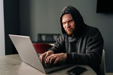 Portrait Of Funny Hacker Male In Black Hoodie Smiling Looking At Camera While Typing On Laptop