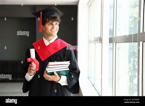 Male Graduate Student With Diploma And Books Near Window In Room Stock
