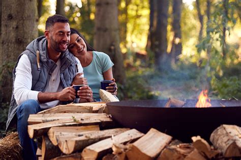 Romantic Couple Camping Sitting By Bonfire In Fire Bowl With Hot Drinks