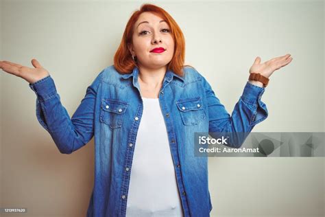 흰색 고립 된 배경 단서와 팔과 손을 들고 혼란 스러운 표정 위에 서 데님 셔츠를 입고 젊은 아름 다운 빨간 머리 여자 의심