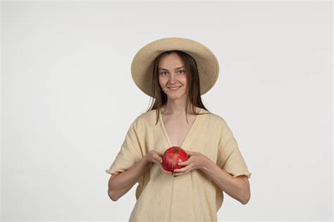밝은 스튜디오에서 긴 머리의 젊은 여성이 빨간 사과를 양손으로 들고 미소를 짓고 있습니다 밝은 색의 의상과 밀짚모자를 착용하고 쾌활한 분위기를 자아내고 있습니다 30 34