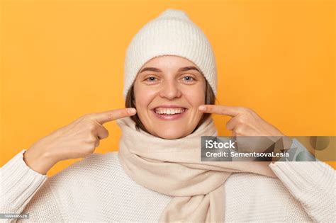 흰색 스웨터 모자 스카프를 착용하고 검지 손가락으로 이빨 미소를 가리키며 미백이나 치과 의사 방문 후 잘 관리된 하얀 치아를
