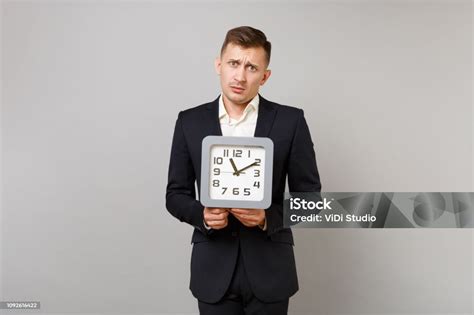 고전적인 검은 양복과 셔츠 스튜디오에 회색 벽 배경에 고립 된 사각형 시계를 들고 의아해 젊은 사업가의 초상화 성과 경력 부 비즈니스 개념입니다 복사 공간을 비웃는 다 검은