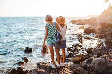 힙스터를 입은 두 명의 젊은 쾌활한 여성이 바다 해안의 바위 위에 모자를 씁니다 소녀 바다 섬 및 오렌지 햇빛이있는 여름 풍경 20 24세에 대한 스톡 사진 및 기타 이미지