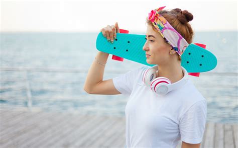 그녀의 머리에 두건을 가진 세련된 젊은 여성 해변에 그녀의 손에 스케이트 보드를 들고 헤드폰 복고풍 스타일 대중 문화 80 년대 청소년 개념 T 셔츠에 대한 스톡 사진 및