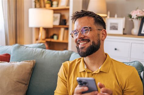 거실 소파에 앉아 스마트폰을 사용하고 있는 중년의 쾌활하고 꽃미남 남자 30 34세에 대한 스톡 사진 및 기타 이미지 30 34세 Brand Name Smart