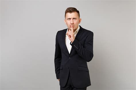 엄격한 젊은 사업가 자장을 말하는 고전적인 검은 양복 셔츠에 회색 배경에 고립 입술 쉿 제스처에 손가락으로 조용할 성과 경력 부