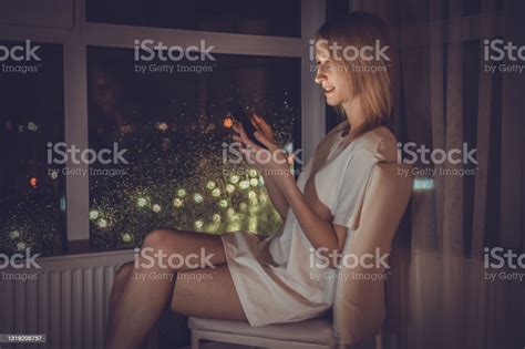 그녀의 손에 휴대 전화와 흰색 셔츠를 입은 아름다운 젊은 금발 의자는 바닥에서 천장까지 의자 및 야간 도시 근처의 의자에 앉아
