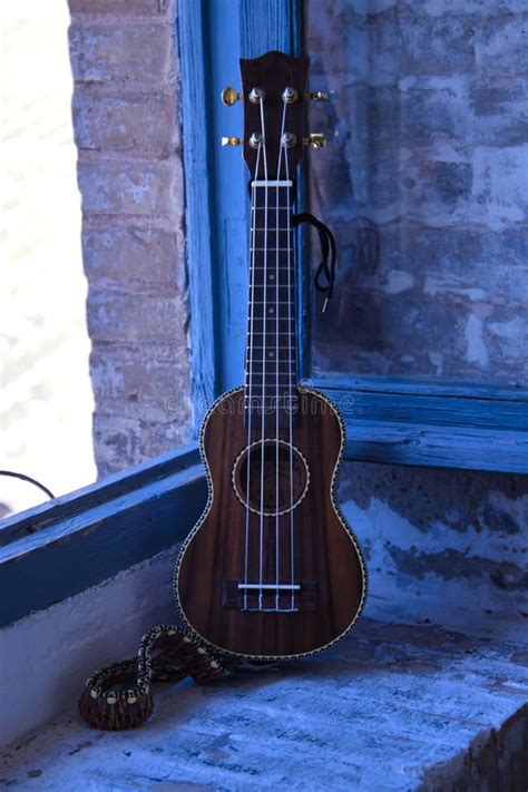 Brown Colored Ukulele With Strap Leaning Against A Brick Window Frame Concept Instruments