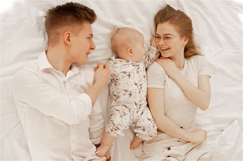 젊은 어머니와 아버지는 침대에 작은 아기와 함께 누워 재미 있고 흰색 배경에서 즐겁습니다 부모와 유아 사이의 귀여운 관계 무료