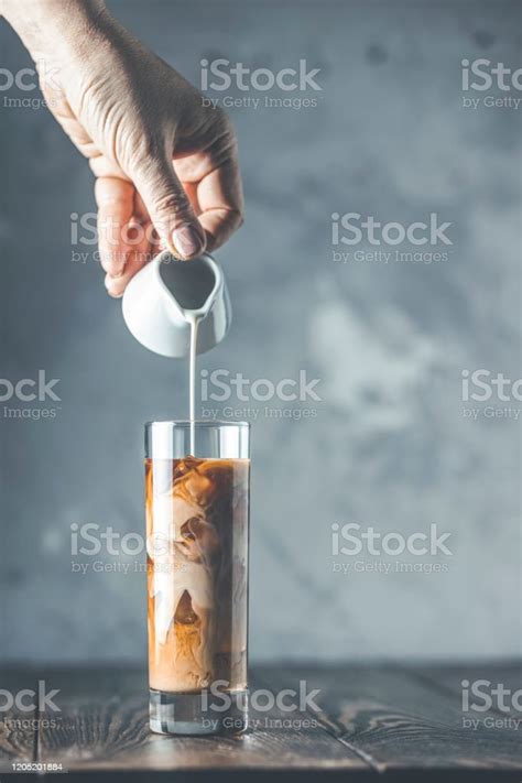 여성의 손은 차가운 커피와 얼음을 양조 유리에 작은 항아리에서 수제 사워 크림을 붓고있다 어두운 나무 테이블에 차가운 여름