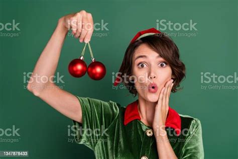 깜짝 놀란 충격적인 아가씨의 사진은 두 개의 Baubles 오픈 입을 착용 엘프 의상 모자 고립 된 녹색 색상 배경을 개최 갈색