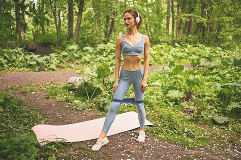 핑크 요가 매트와 여름 녹색 공원에서 피트니스 트레이닝 껌과 함께 스쿼트 운동을하고 큰 흰색 헤드폰과 블루 스포츠웨어에 젊은 아름다운 슬림 핏 소녀 완벽한 몸을 가진 소녀