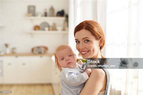 매력적인 매력적인 젊은 엄마의 실내 이미지 생강 머리와 주 근 깨 간호 그녀의 귀여운 아기 잠 그를 노래 그녀의 팔에 유아와